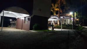 Cena noturna de um edifício com área externa coberta, calçada pavimentada e pequeno jardim com uma estátua.