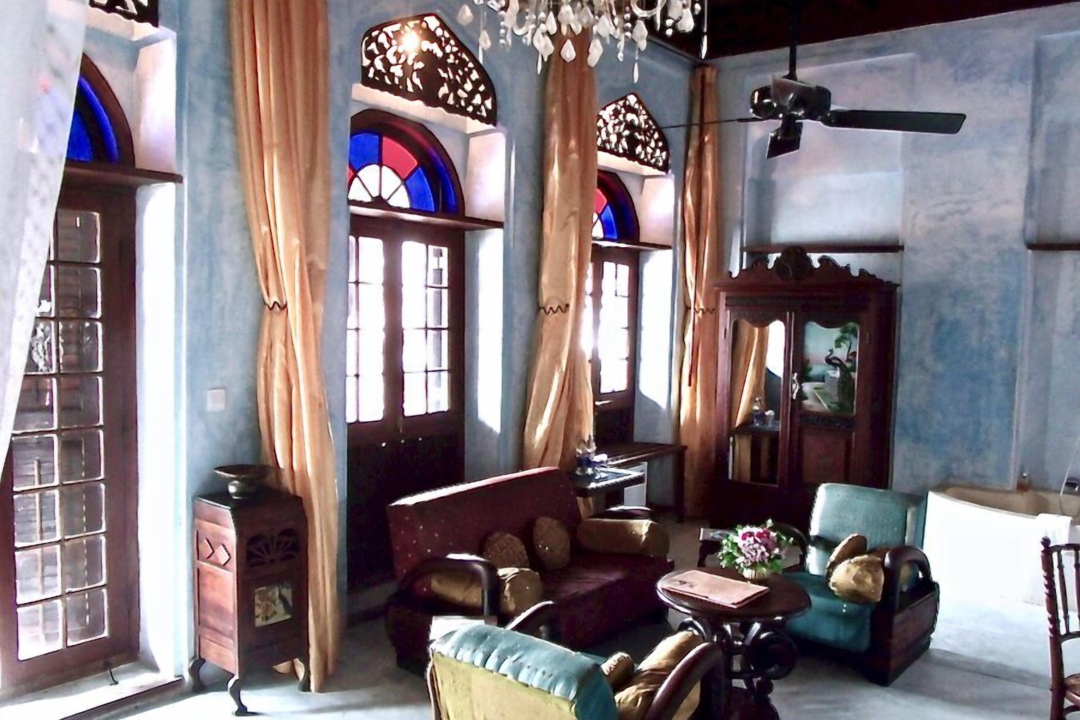 Quarto com sofá de madeira, uma pequena mesa e uma cadeira. Há cortinas nas janelas, e um ventilador de teto é visível.