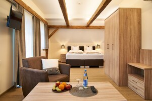 Quarto de hotel com teto de madeira, uma cama com roupa de cama branca, uma poltrona marrom, uma mesa pequena com frutas e uma garrafa, e uma TV presa na parede.