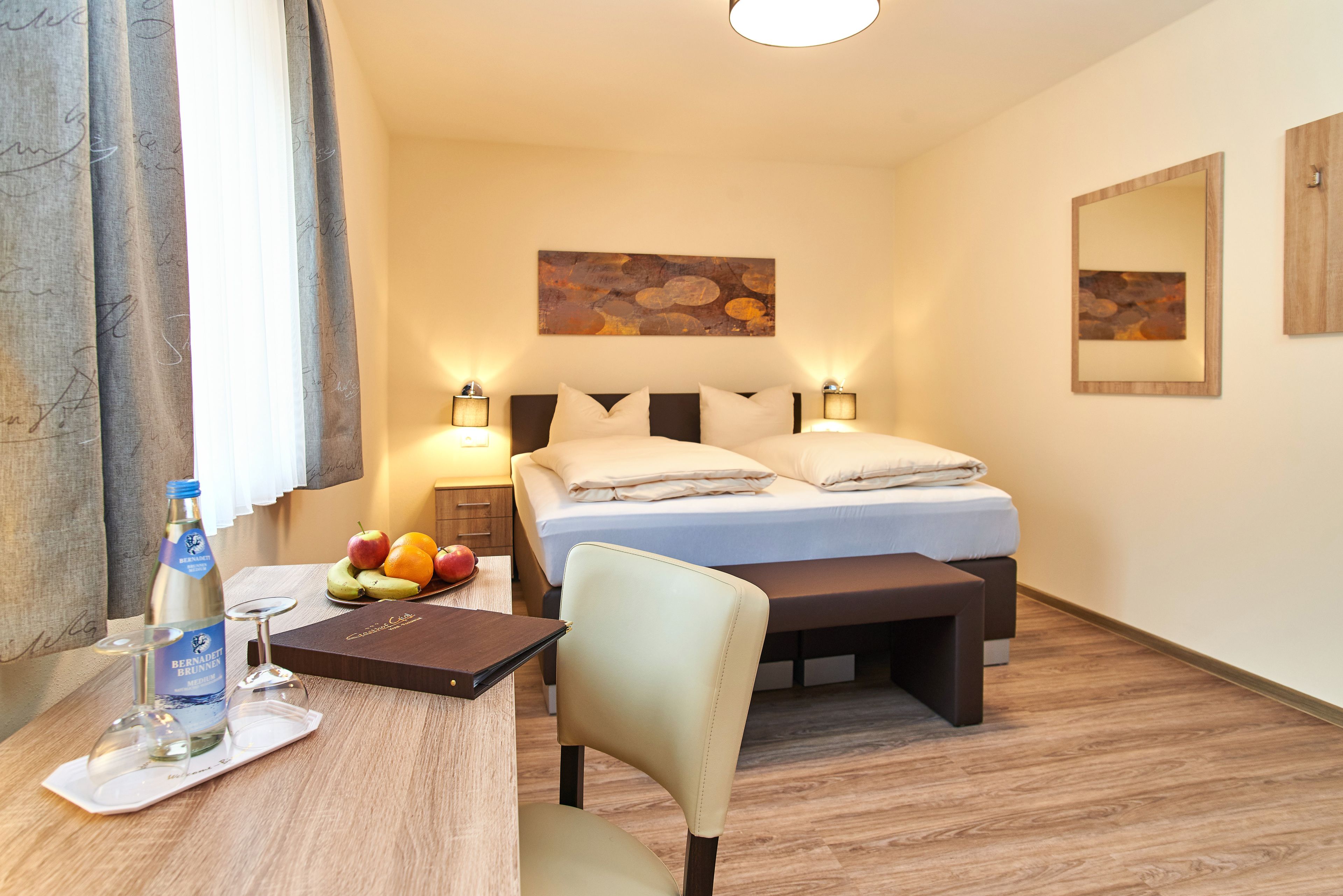 Quarto de hotel com cama, luminárias de cabeceira, escrivaninha com garrafa e copos, uma cadeira e uma tigela de frutas.
