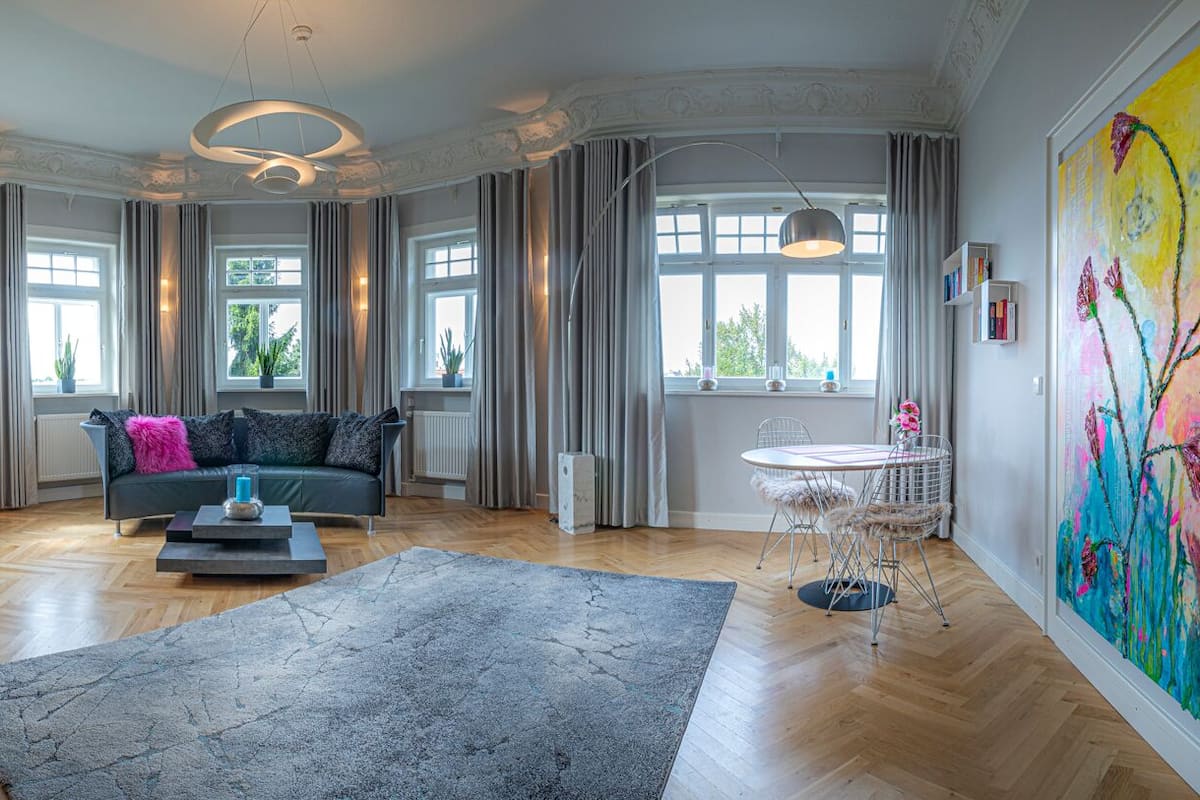 Uma sala de estar ampla com um sofá moderno, uma mesa de centro e um espaço de jantar com uma mesa pequena e cadeiras. Há janelas grandes com cortinas, um quadro na parede e um lustre.