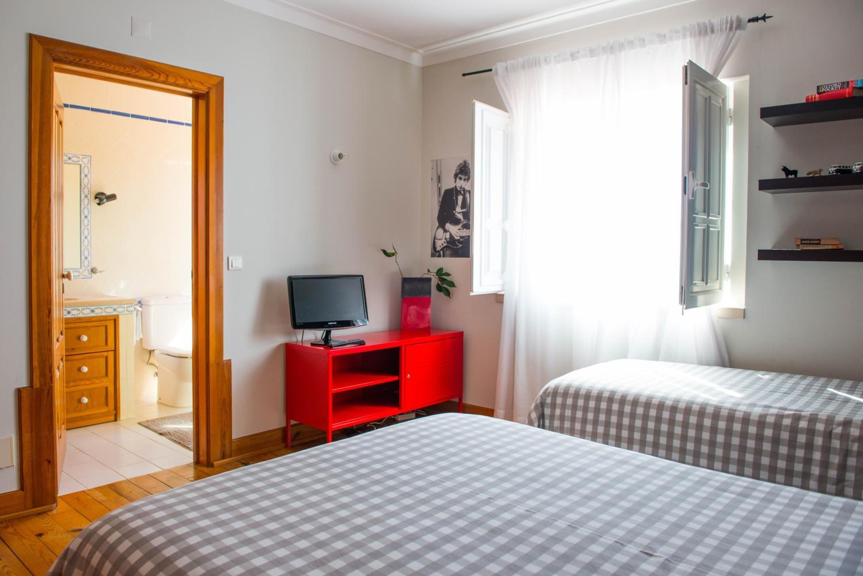 Quarto com duas camas, uma TV sobre um suporte vermelho, uma janela com cortinas e uma estante com livros.