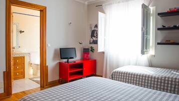 Quarto com duas camas, uma TV sobre um suporte vermelho, uma janela com cortinas e uma estante com livros.