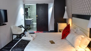 Quarto de hotel com cama, poltrona, televisão e banheiro visível através de uma porta aberta.