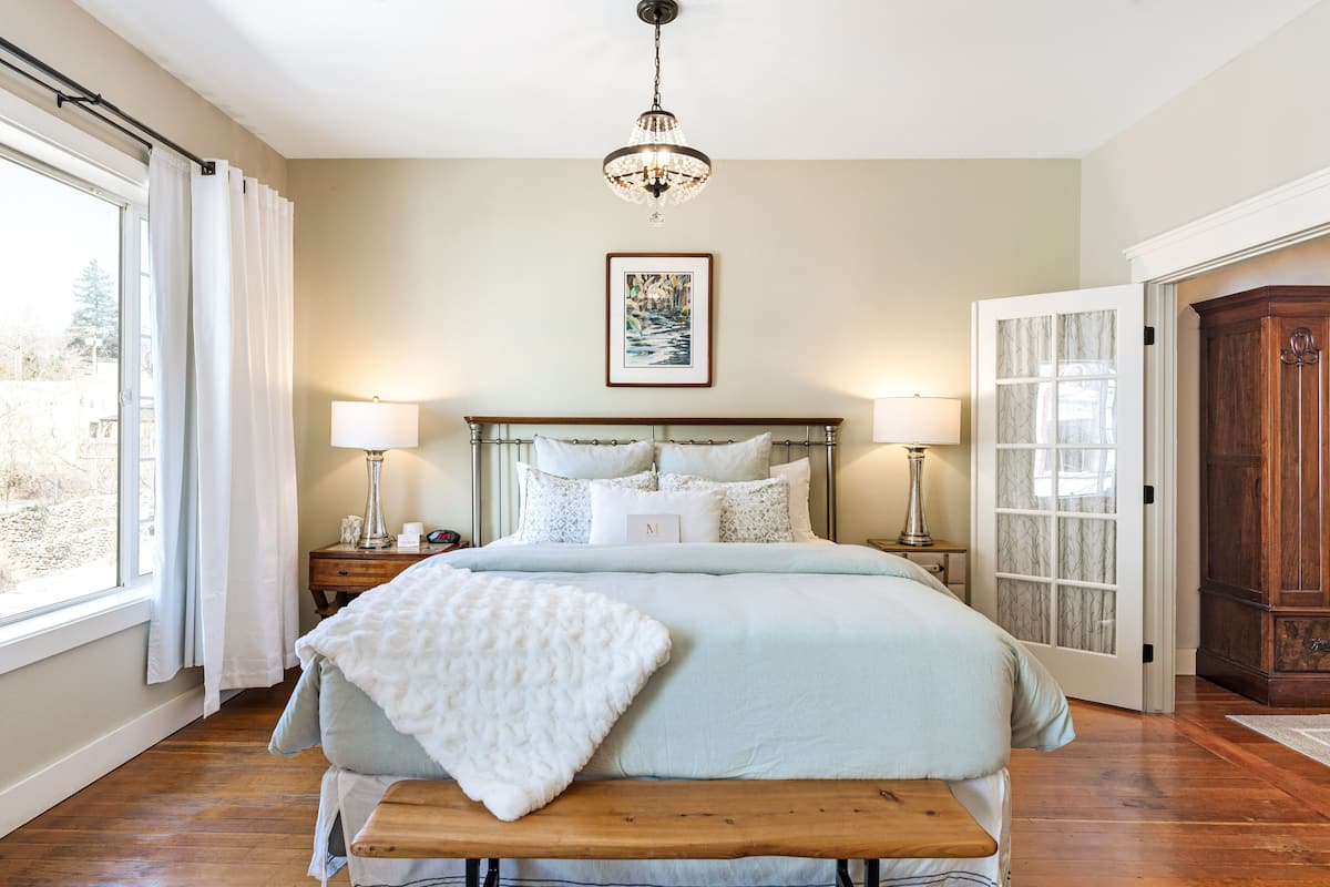 Quarto com cama grande, duas luminárias de cabeceira, um lustre, uma moldura na parede e um banco de madeira.