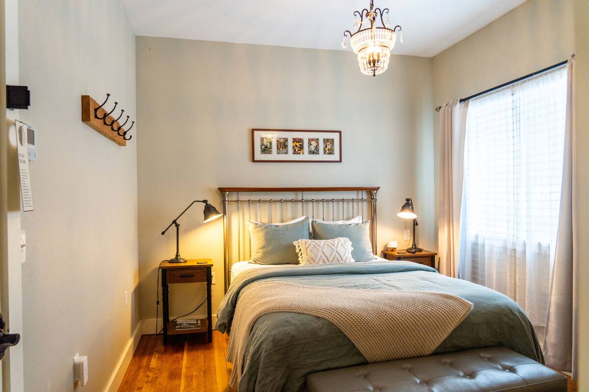 Quarto com cama, luminárias de cabeceira, um candelabro, um quadro na parede e uma janela com cortinas.