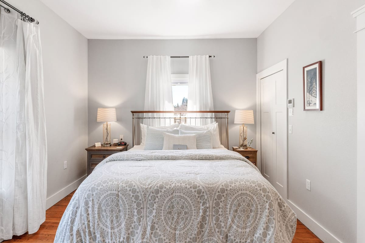 Quarto com uma cama grande, duas mesas de cabeceira com luminárias, uma janela com cortinas leves e uma porta com um quadro emoldurado.