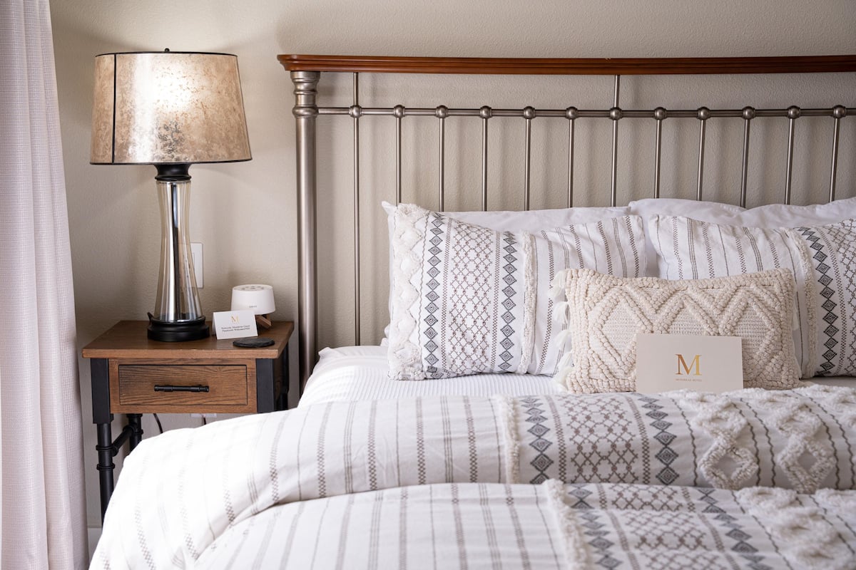 Cama bem arrumada com roupa de cama estampada, luminária de cabeceira, mesa de cabeceira pequena de madeira com gaveta e um cartão com nome na cama.