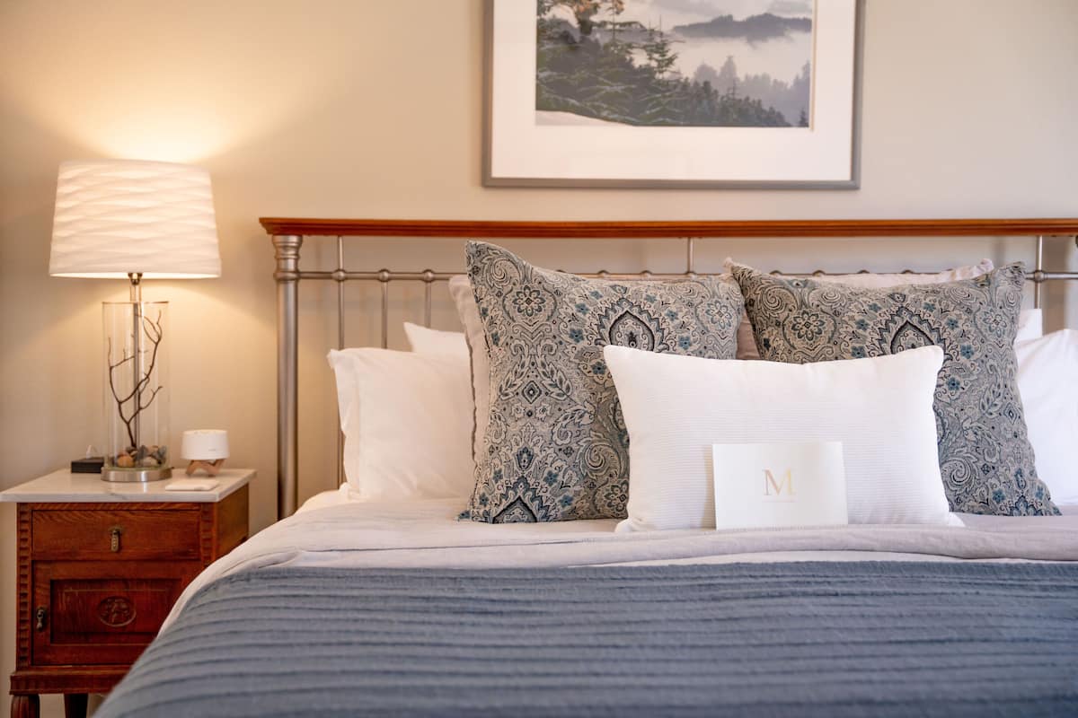 Cama bem arrumada com almofadas estampadas e uma paisagem emoldurada acima.