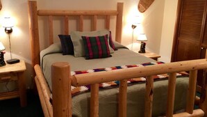 Uma cama de madeira com cabeceira, duas mesas de cabeceira com abajures e um cobertor estampado.