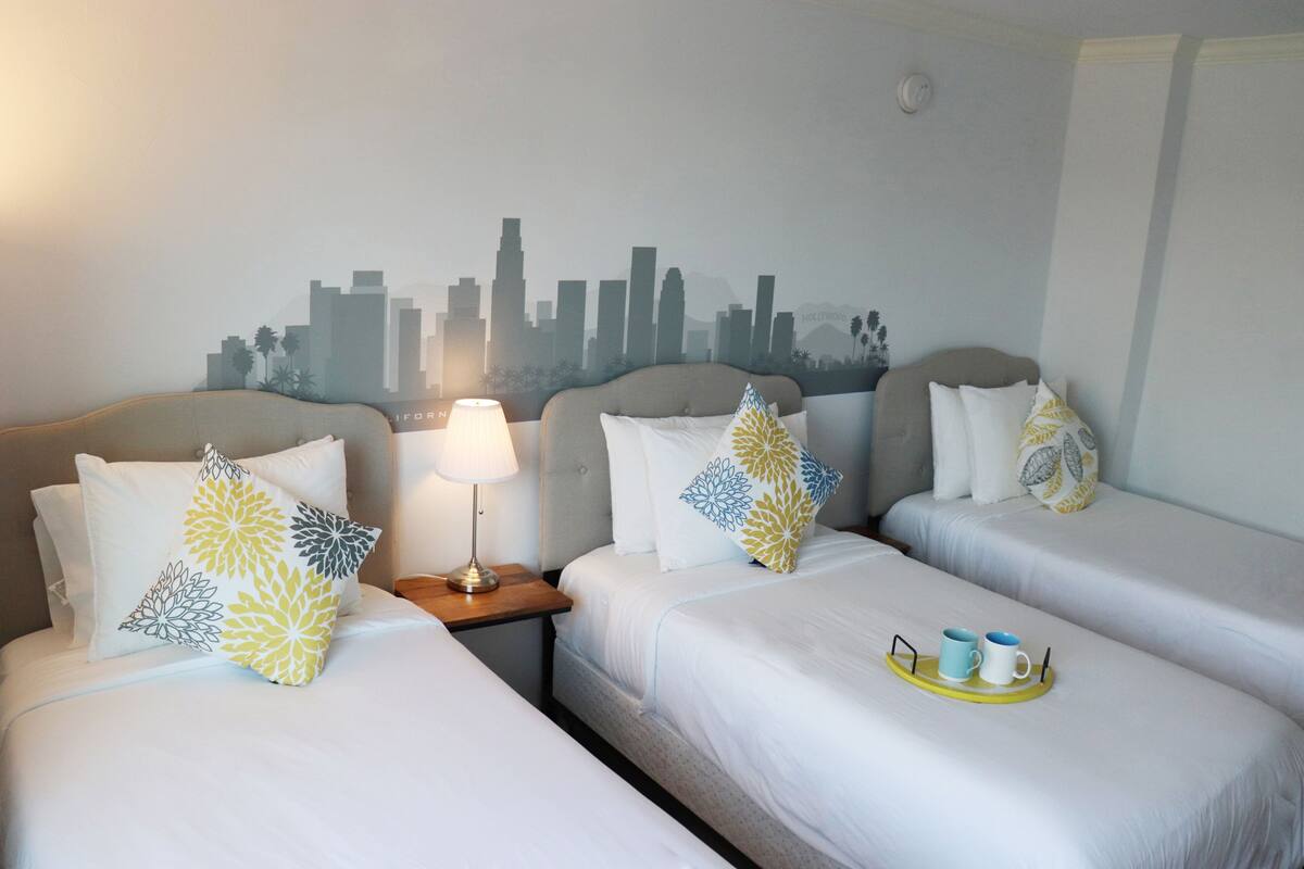 Quarto de hotel com três camas, um mural da paisagem urbana na parede, uma mesinha de cabeceira com abajur e um jogo de chá com duas xícaras.