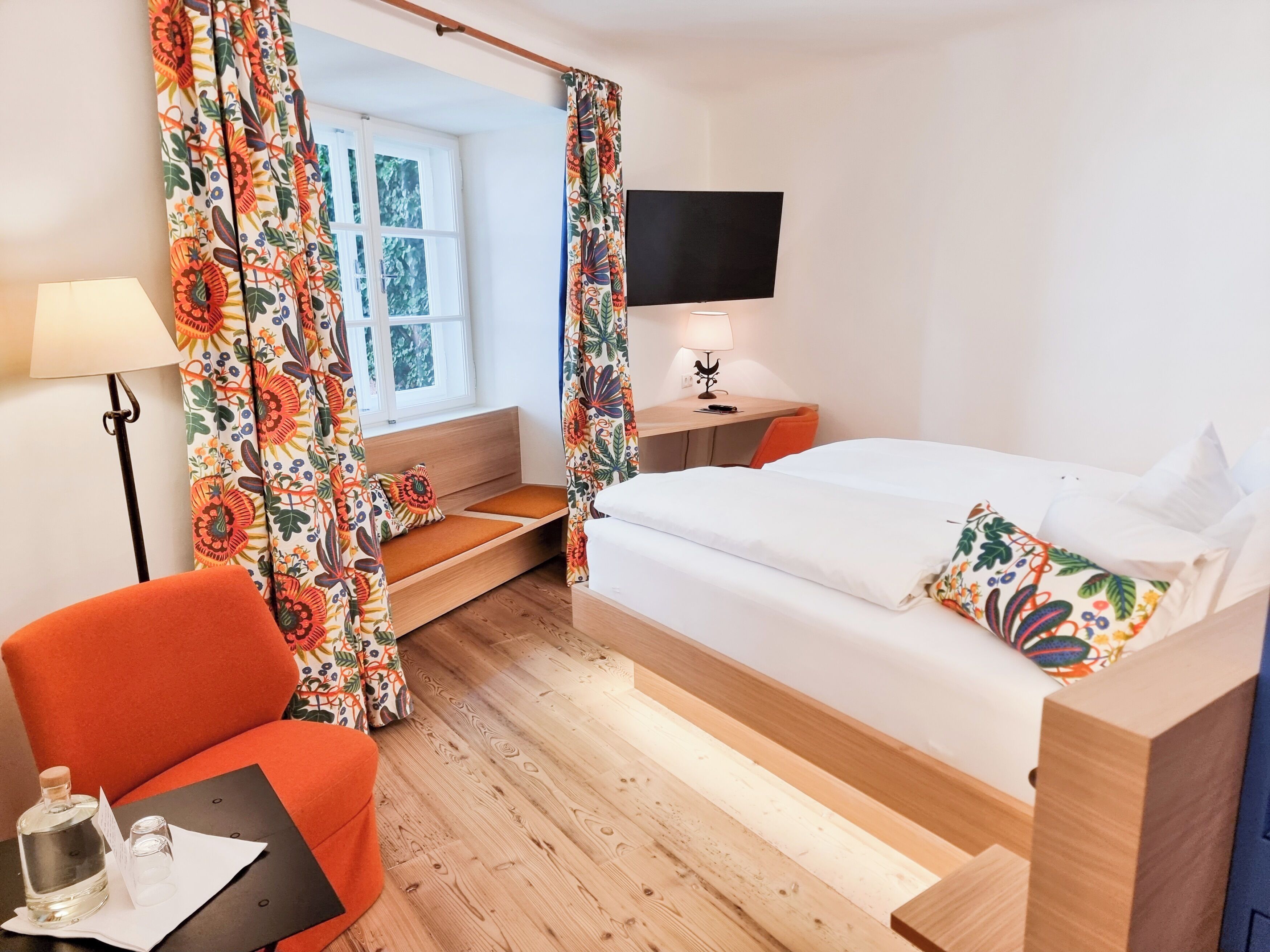 Quarto de hotel com cama, escrivaninha, cadeira, televisão e janela com cortinas.