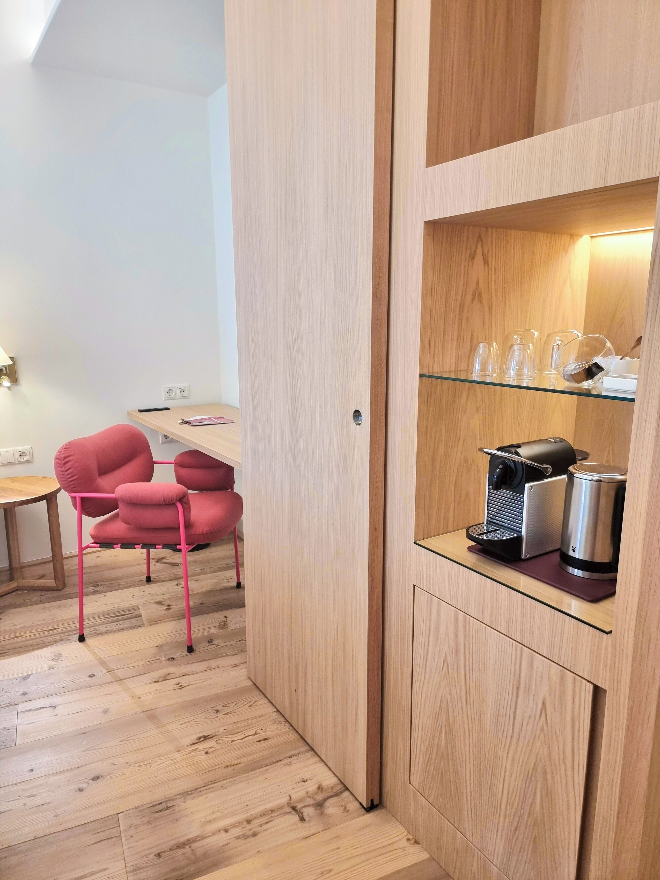 Quarto compacto com piso de madeira, uma cadeira rosa, uma pequena mesa e prateleira de vidro com eletrodomésticos.