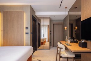 Quarto de hotel com cama, escrivaninha com cadeira, televisão e espelho.