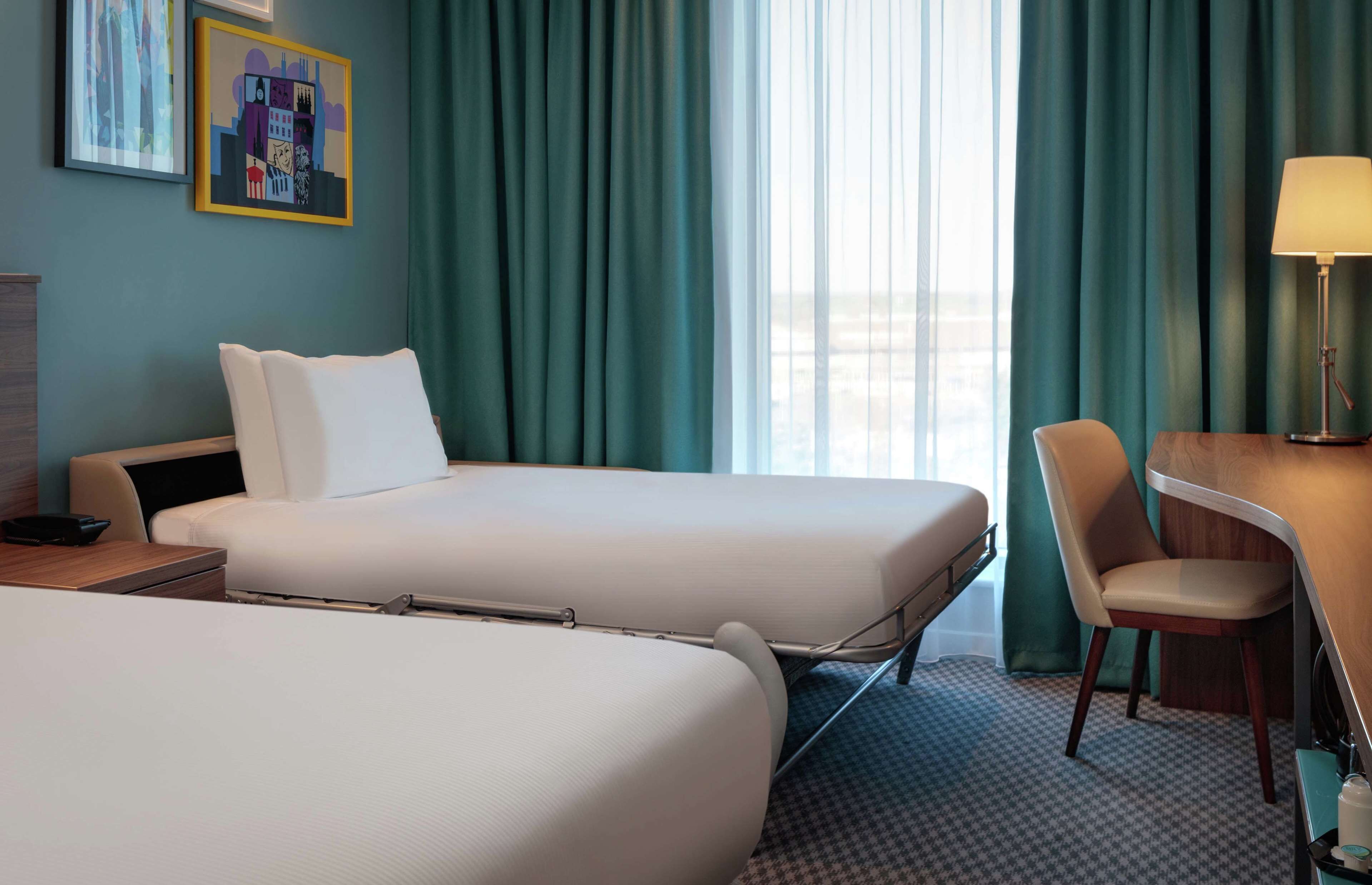 Quarto de hotel com duas camas, uma escrivaninha, uma cadeira, um abajur e vista para a cidade pela janela.