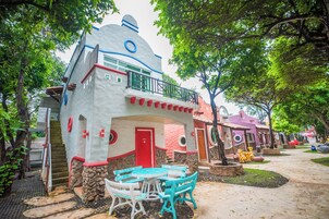 Um edifício singular com porta vermelha, varanda e pequena área externa com mesas e cadeiras.