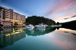 Um hotel à beira-mar com várias varandas, barcos atracados na água e uma colina arborizada ao fundo.