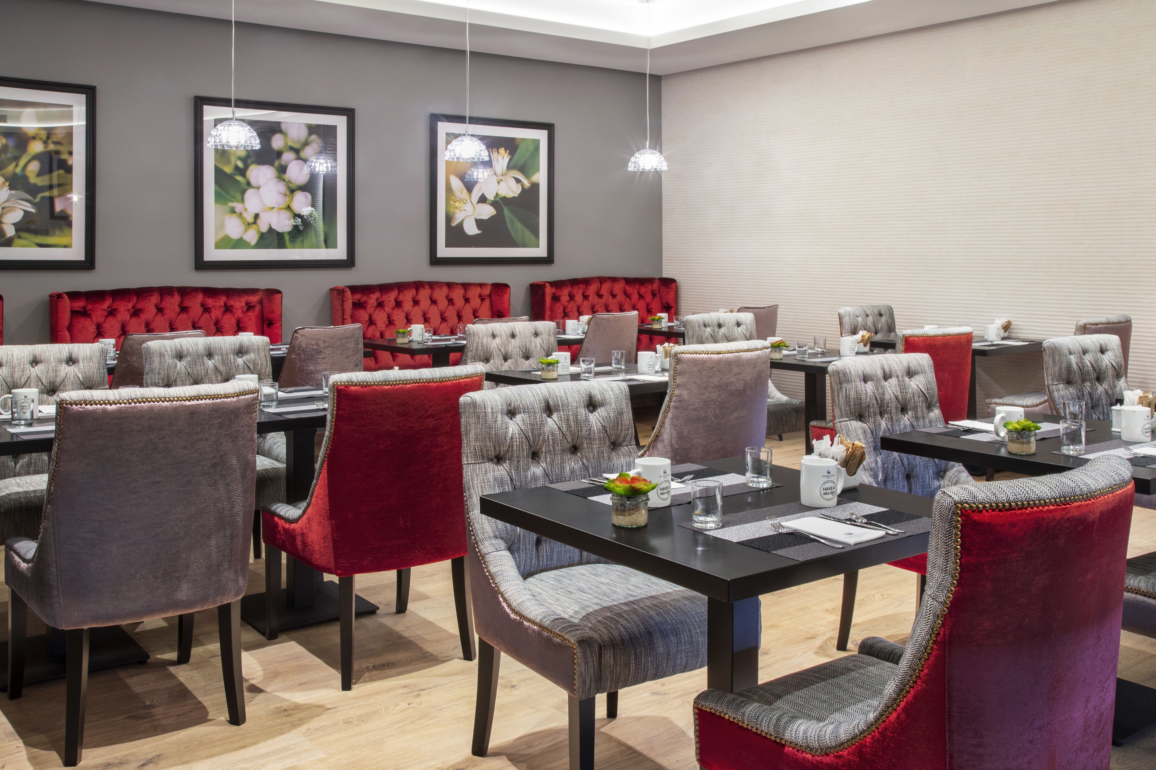 Uma sala de jantar de hotel com bancos estofados em vermelho e cadeiras estofadas em cinza, dispostas ao redor de mesas pretas com jogos americanos brancos e talheres.