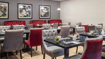 Uma sala de jantar de hotel com bancos estofados em vermelho e cadeiras estofadas em cinza, dispostas ao redor de mesas pretas com jogos americanos brancos e talheres.