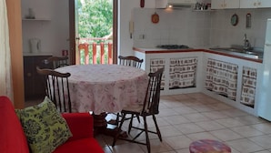 Uma sala de estar aconchegante com um sofá vermelho, uma mesa de centro redonda e uma área de jantar com mesa e cadeiras. Uma cozinha com armários brancos e um fogão a gás é visível ao fundo.