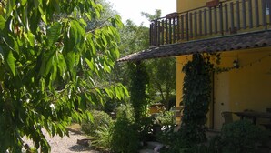 Um edifício amarelo com varanda, rodeado por vegetação e um caminho de cascalho.