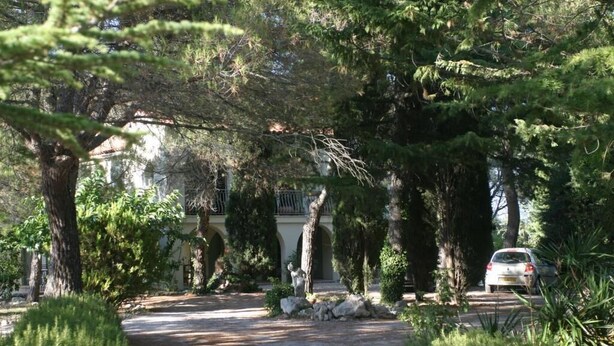 Uma entrada de automóveis leva a um edifício com fachada de pedra e janelas em arco, cercado por árvores grandes e um carro estacionado.