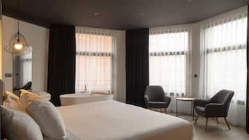 Quarto de hotel moderno com uma cama grande, duas poltronas, uma mesa pequena e uma luminária pendente.