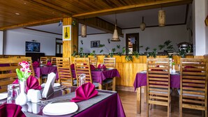 Restaurante com mesas e cadeiras de madeira, toalhas de mesa roxas e luminárias pendentes.