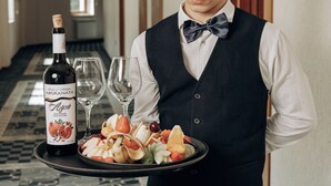Uma pessoa vestindo um colete preto e gravata borboleta carregando uma bandeja com uma garrafa de vinho, copos e uma travessa de frutas.