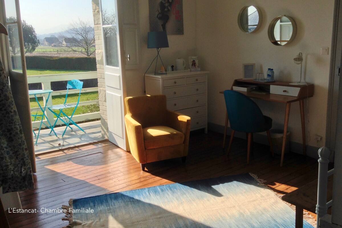 Um quarto com piso de madeira, uma cadeira azul, uma poltrona amarela, uma escrivaninha com computador e uma janela com vista para uma paisagem verde.