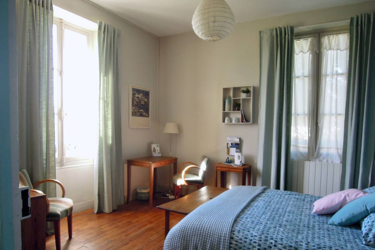 Quarto com cama, cadeira, mesinha, abajur e prateleira com objetos decorativos.