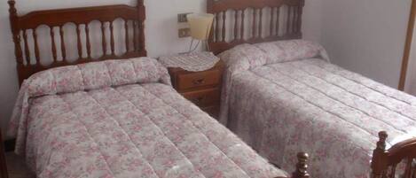 Quarto com duas camas de madeira, cada uma com uma colcha florida. Há uma mesa de cabeceira com abajur entre as camas.