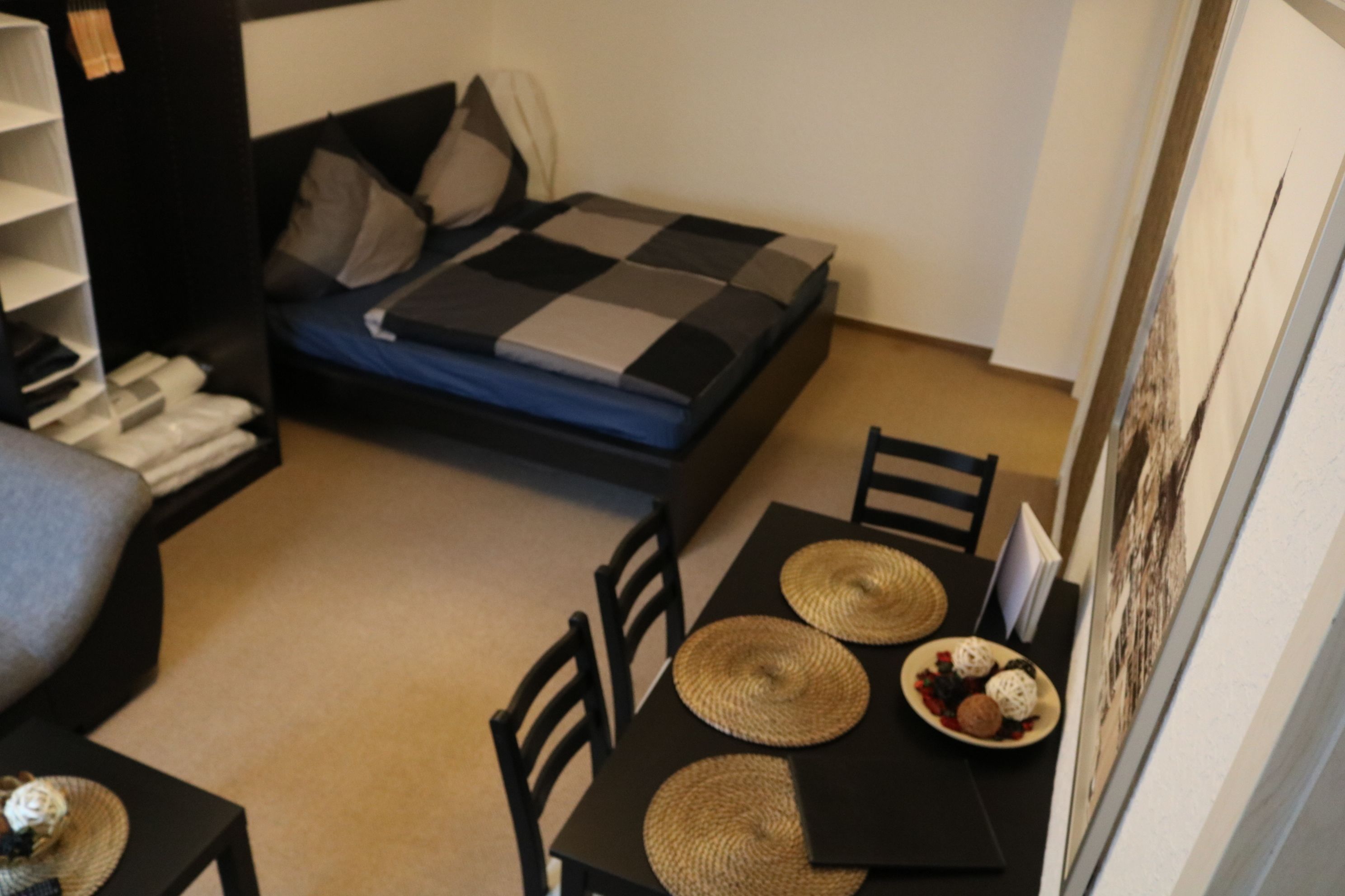 Quarto compacto com cama, mesa de jantar e cadeiras, e um pequeno armário com roupas dobradas.