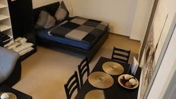 Quarto compacto com cama, mesa de jantar e cadeiras, e um pequeno armário com roupas dobradas.