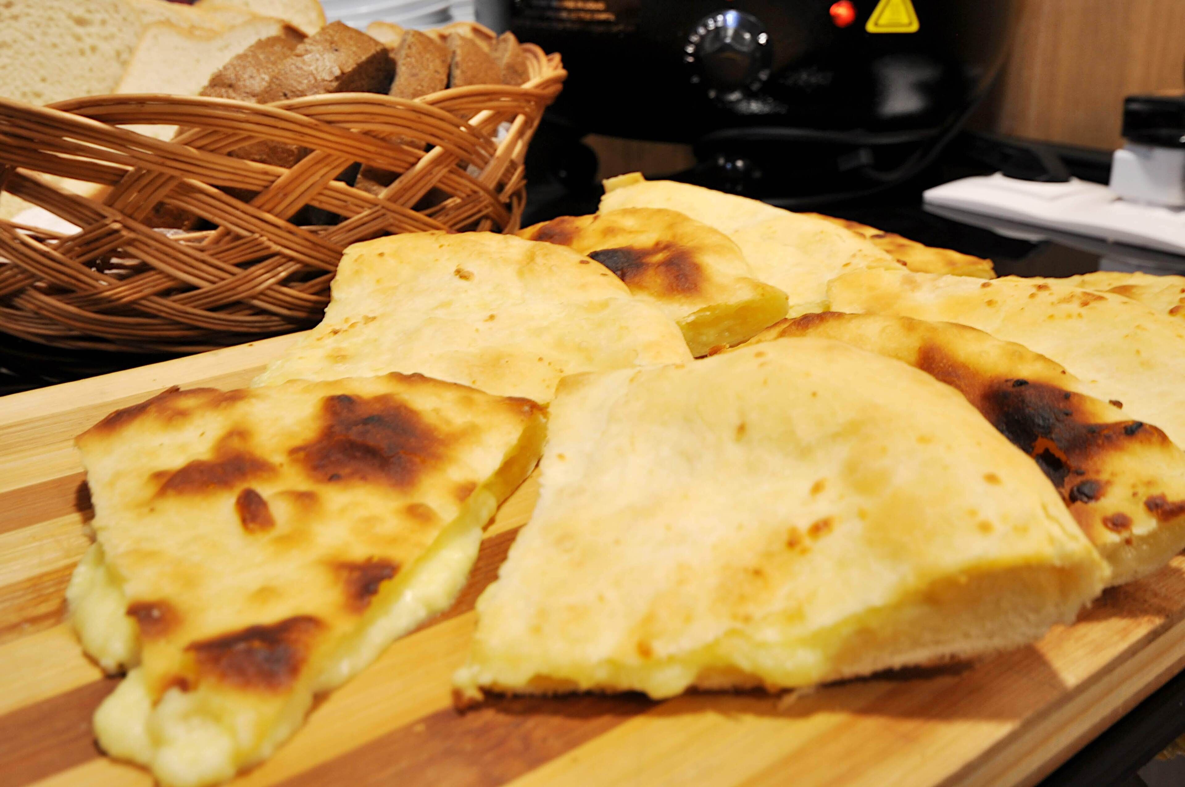 Uma tábua de madeira com vários pedaços de pão achatado, uma cesta de vime e uma máquina de café ao fundo.