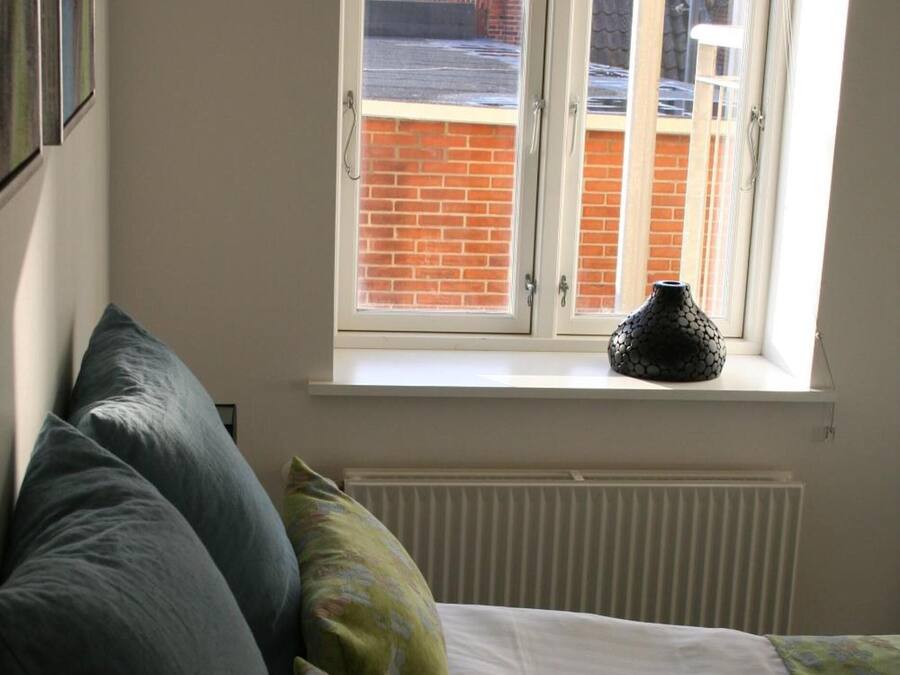 Quarto com cama, janela com vista para um edifício de tijolos e radiador abaixo da janela.