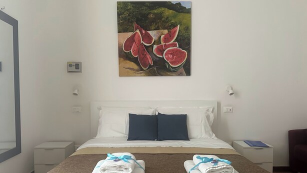 Quarto de hotel com uma cama grande, duas almofadas, uma pintura de melancias e duas toalhas dobradas na cama.