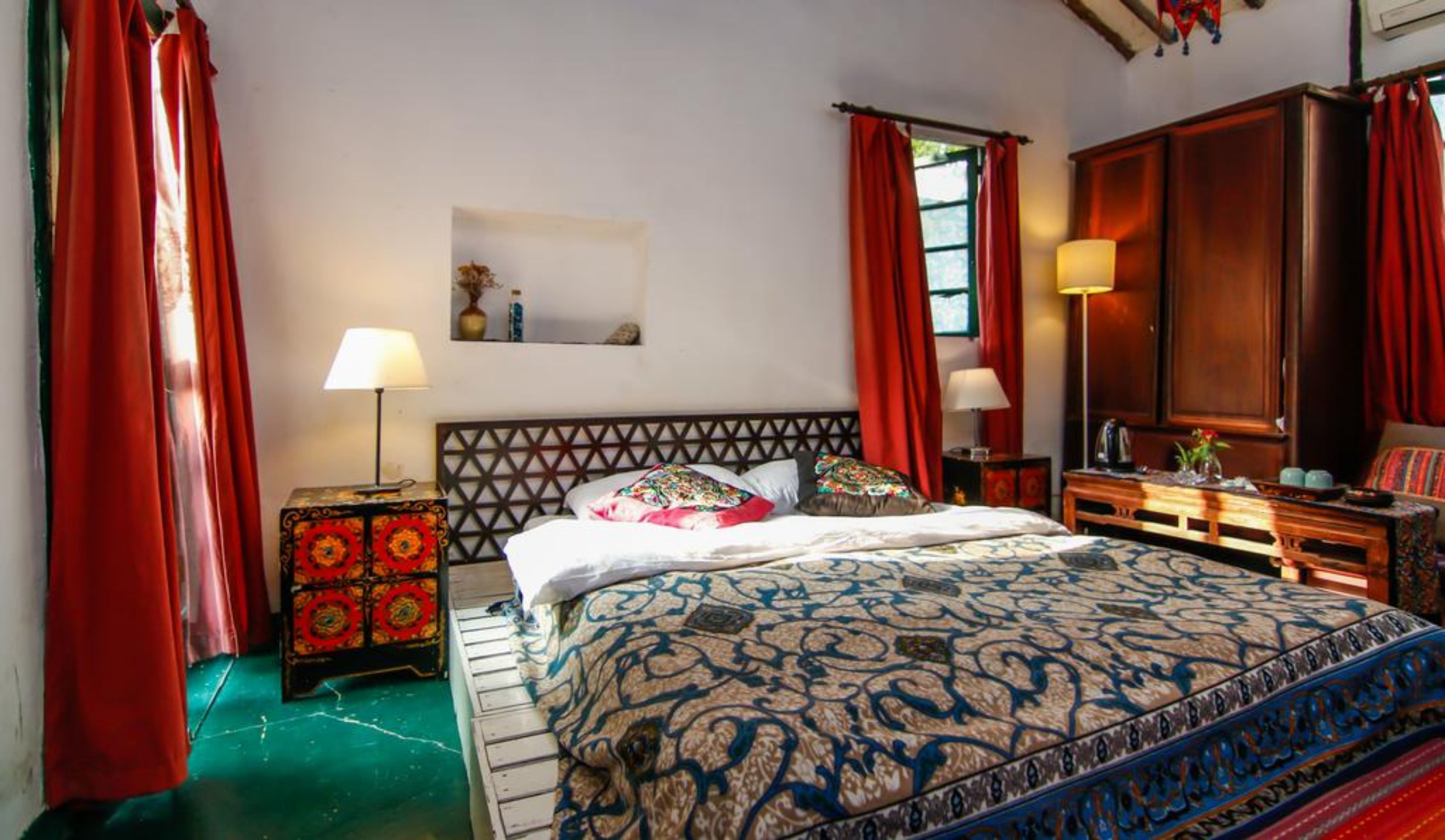 Quarto com uma cama grande, cortinas vermelhas, cabeceira de madeira, mesa de cabeceira com abajur e guarda-roupa de madeira.