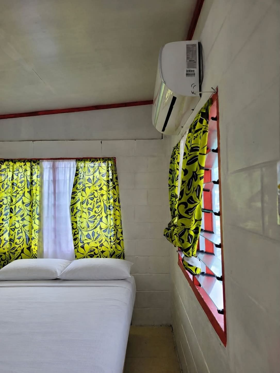 Um quarto pequeno e estreito com uma cama, uma janela com cortinas estampadas e um aparelho de ar condicionado fixado na parede.