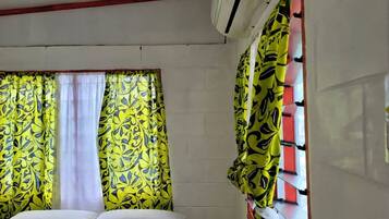 Um quarto pequeno e estreito com uma cama, uma janela com cortinas estampadas e um aparelho de ar condicionado fixado na parede.