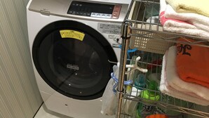 Máquina de lavar com abertura frontal e painel de controle, prateleira de metal com toalhas dobradas e produtos de limpeza.