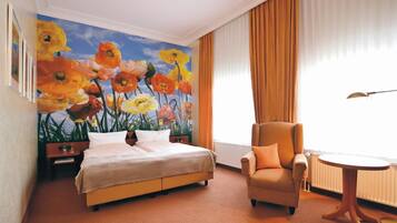Quarto de hotel com cama, uma cadeira, uma mesinha e um quadro de flores na parede.