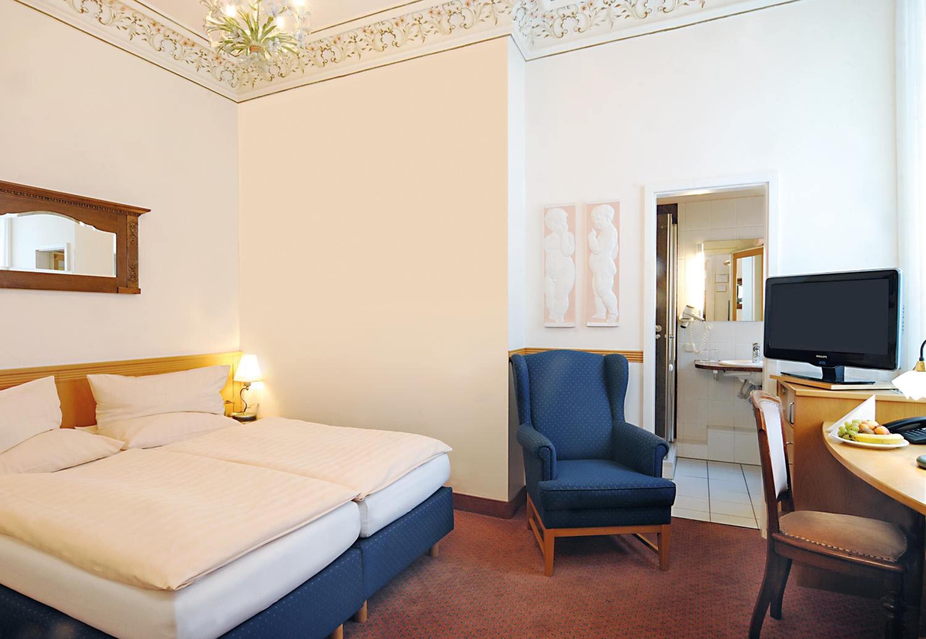 Quarto de hotel com cama, uma cadeira, escrivaninha com computador e televisão.