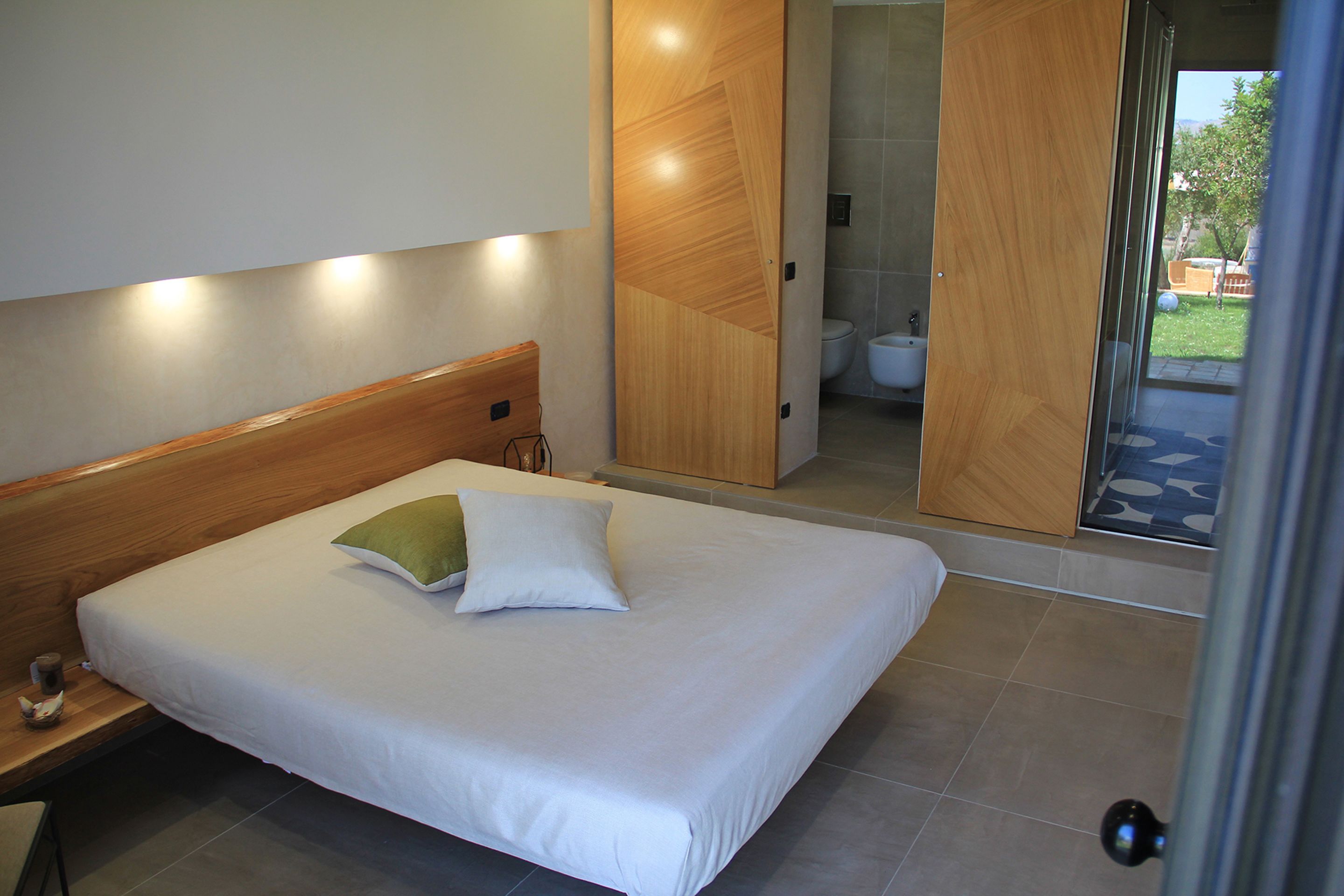 Quarto moderno com cabeceira de madeira, cama com roupa de cama branca e travesseiro verde, mesa de cabeceira e vista para um banheiro e área externa através de uma porta aberta.