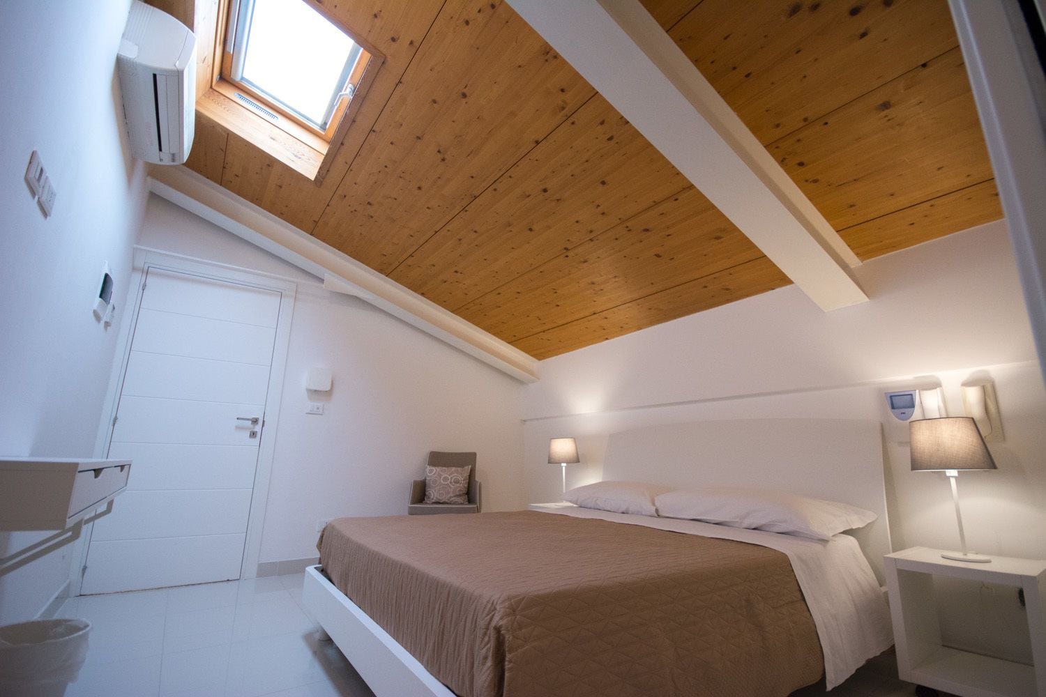 Quarto com teto inclinado, cama com colcha marrom, luminárias de cabeceira, uma cadeira pequena e uma janela.