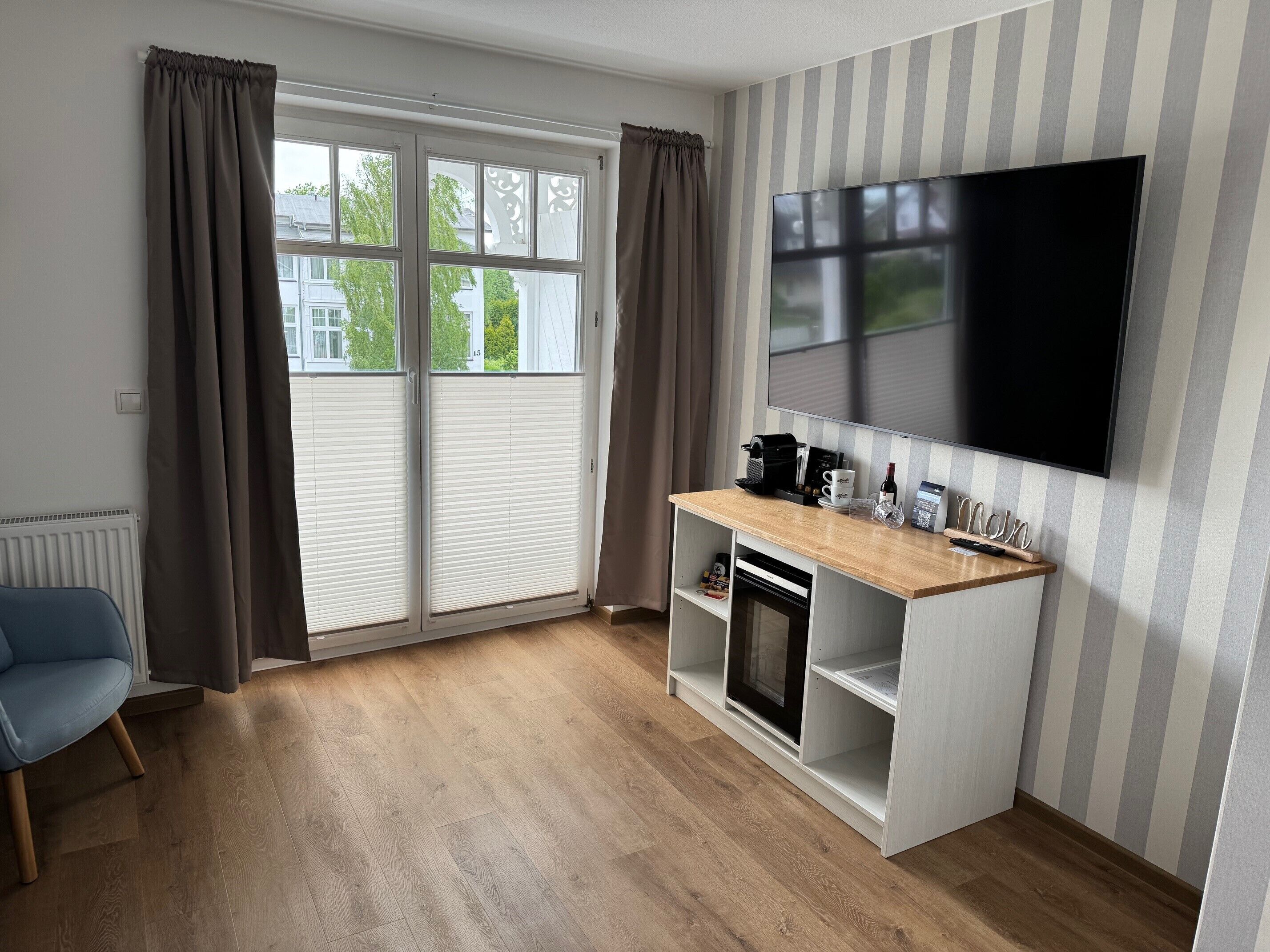 Quarto moderno com uma janela grande, televisão de tela plana, escrivaninha de madeira e uma cadeira azul.