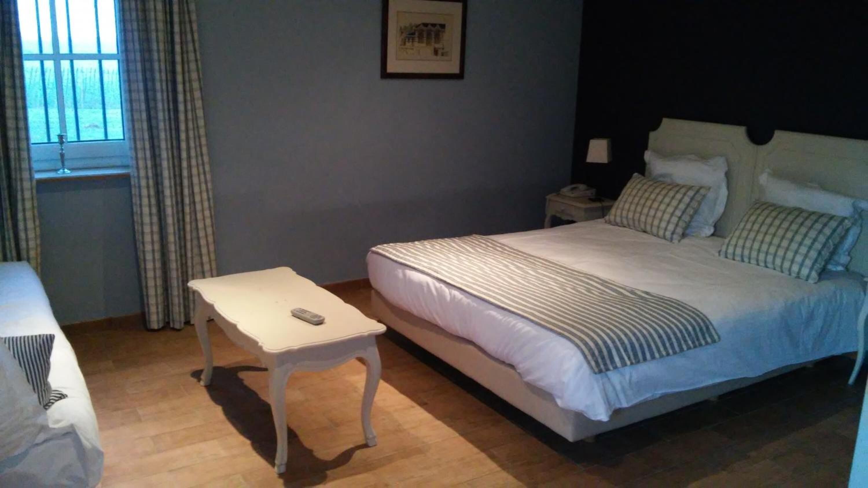 Quarto com cama, mesa de cabeceira, cortinas na janela e um quadro na parede.
