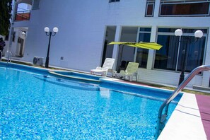 Piscina com água azul cristalina, rodeada por espreguiçadeiras brancas e um guarda-sol amarelo.