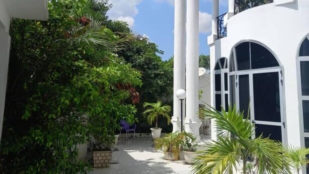 Um edifício branco com colunas e janelas em arco, rodeado por vegetação exuberante e vasos de plantas.