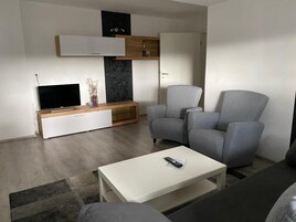 Uma sala de estar moderna com uma televisão de tela plana, uma mesa de centro branca, duas poltronas cinzas e um móvel para aparelhos eletrônicos de madeira.
